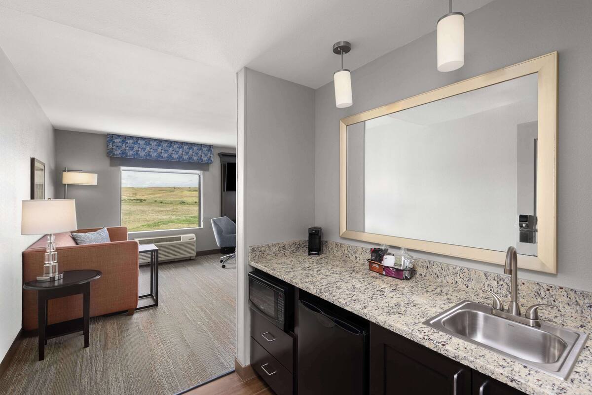 Quarto de hotel compacto com cozinha compacta, um espelho grande, área de refeições com mesa e cadeiras, e vista para o exterior.