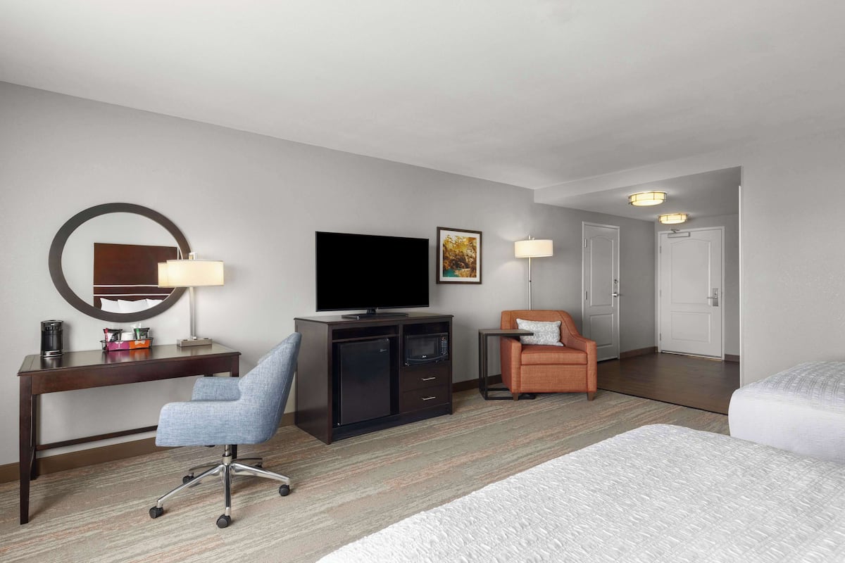 Quarto de hotel com cama, escrivaninha com computador e televisão, uma cadeira, micro-ondas e uma porta.