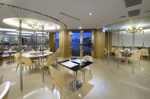 Uma sala de jantar moderna com mesas redondas, cadeiras bege e vista para a cidade através de amplas janelas.