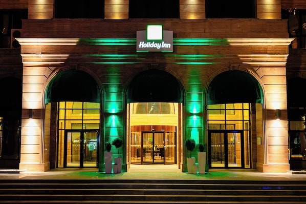 Entrada de um hotel Holiday Inn, com uma placa bem visível, amplas portas de vidro e fachada simétrica com três arcos.