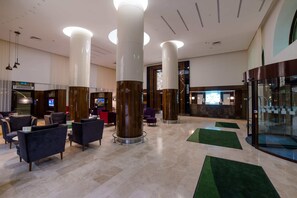 Um hall com grandes colunas cilíndricas, áreas de estar, balcão de recepção e elevador de vidro.