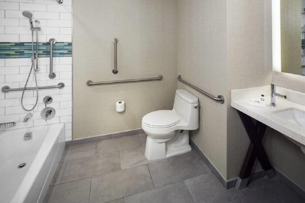 Uma casa de banho moderna com sanita branca, banheira e lavatório. Existem barras de apoio instaladas nas paredes para segurança.