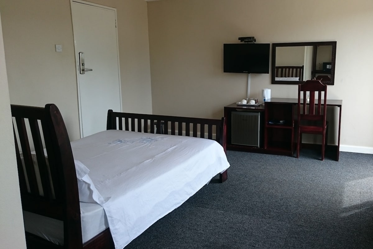 Quarto de hotel com cama, escrivaninha, cadeira, televisão e espelho.
