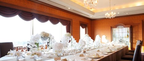 Salão de festas com uma longa mesa posta para um evento formal, com centros de mesa florais brancos e elegantes arranjos de mesa.