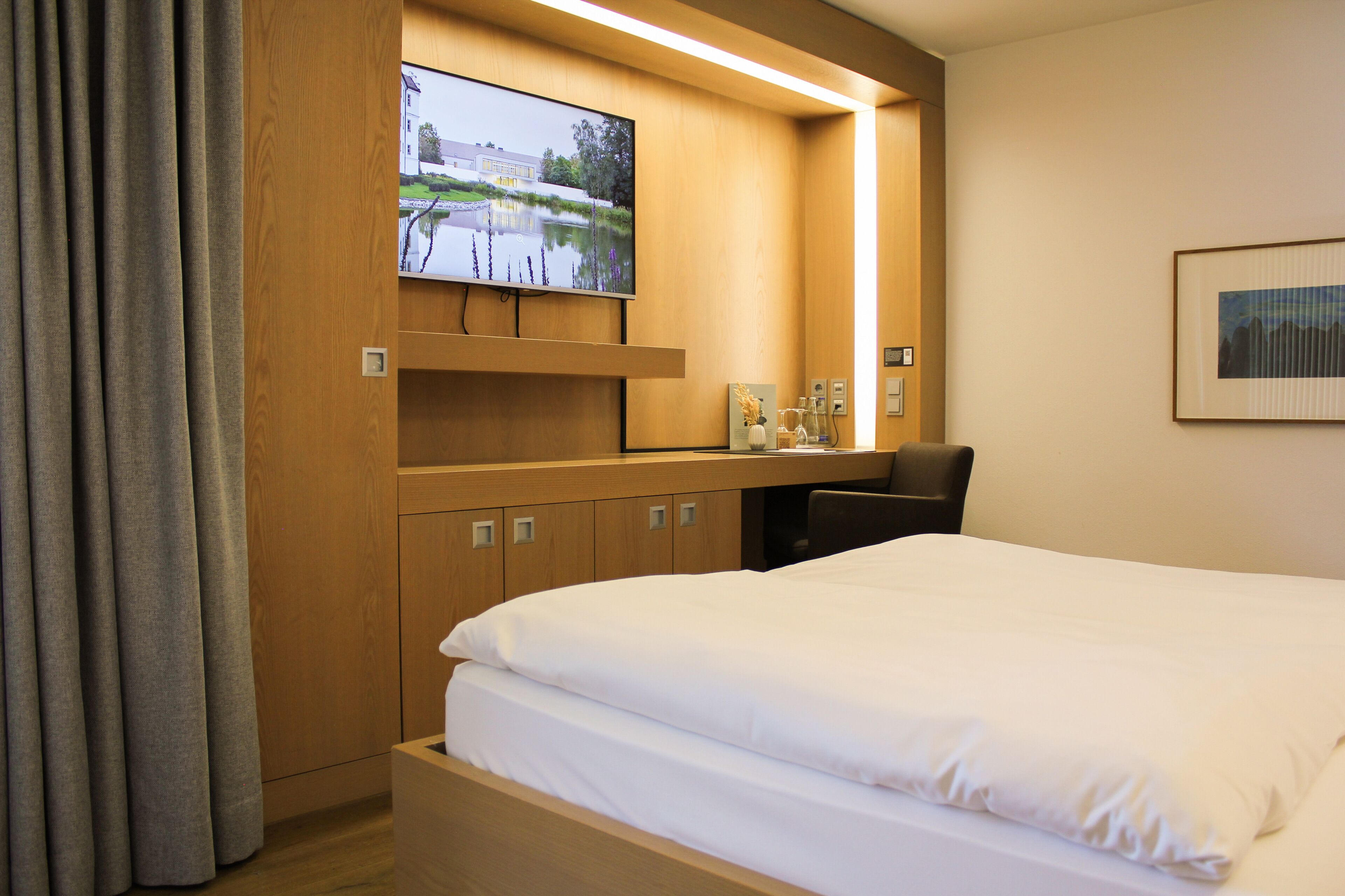 Quarto de hotel com cama, escrivaninha, televisão na parede e um porta-retrato.