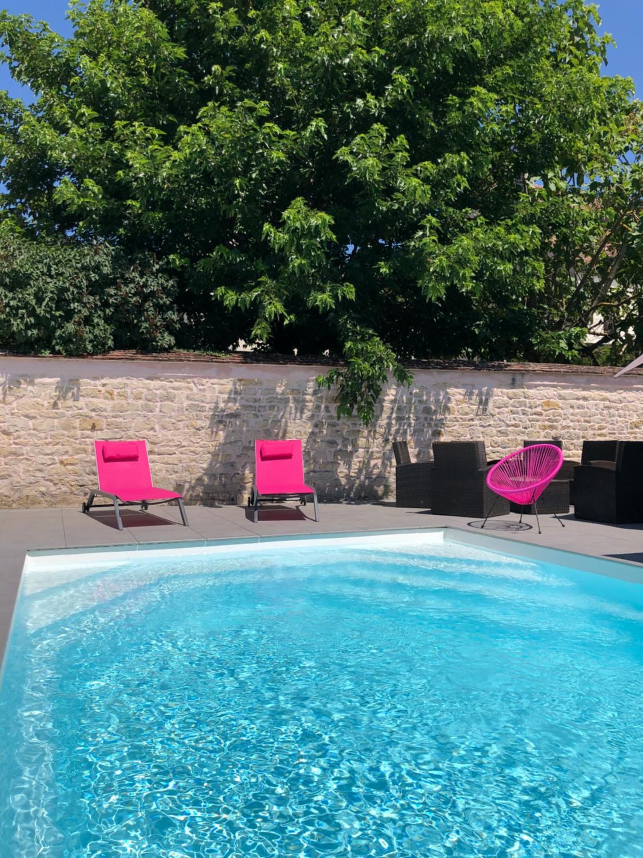 Uma piscina com água azul cristalina, rodeada por um muro de pedra e uma grande árvore. Há duas espreguiçadeiras cor-de-rosa e uma cadeira cor-de-rosa com encosto circular.