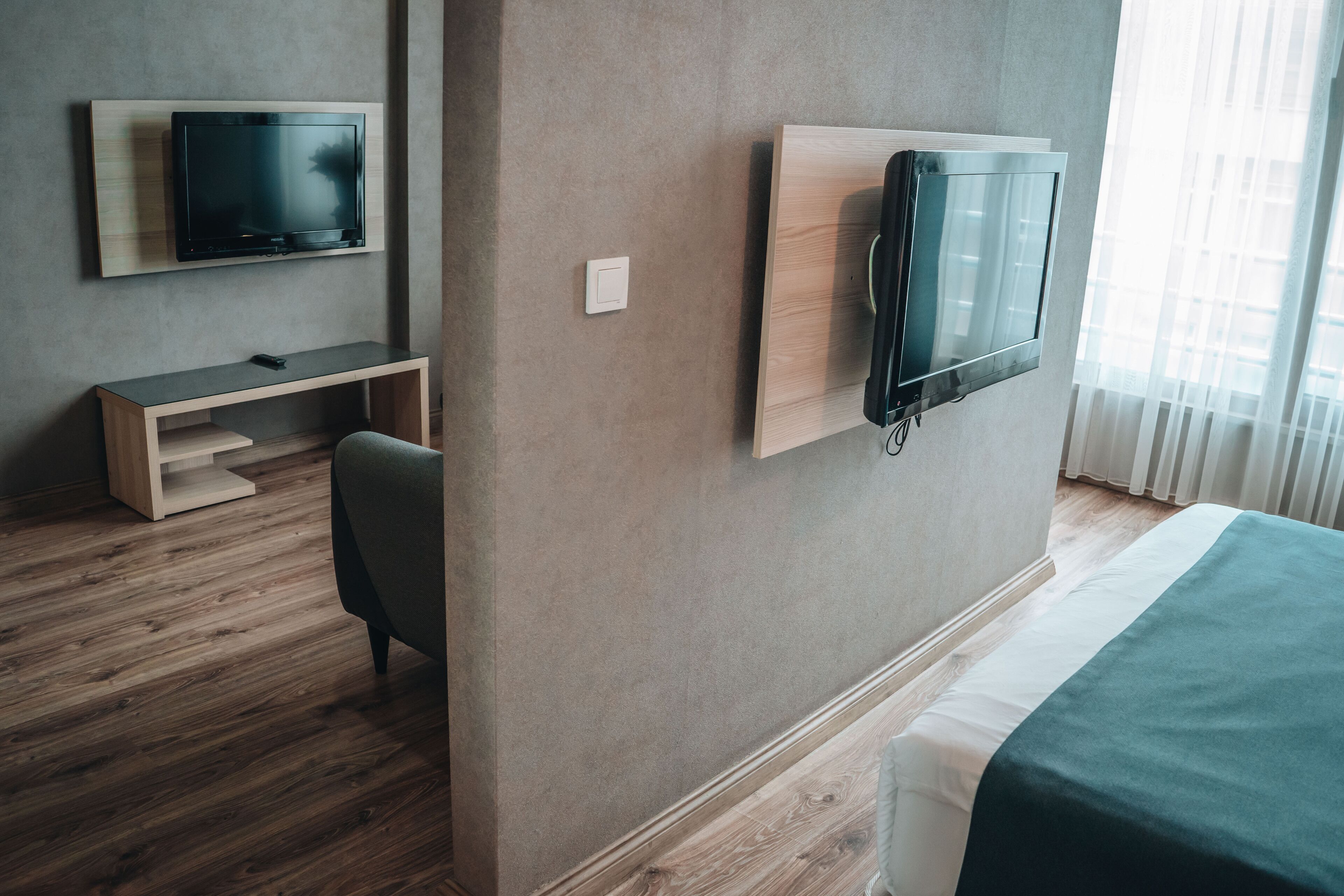 Quarto de hotel com cama, televisão na parede, um banco de madeira e janela com cortinas.