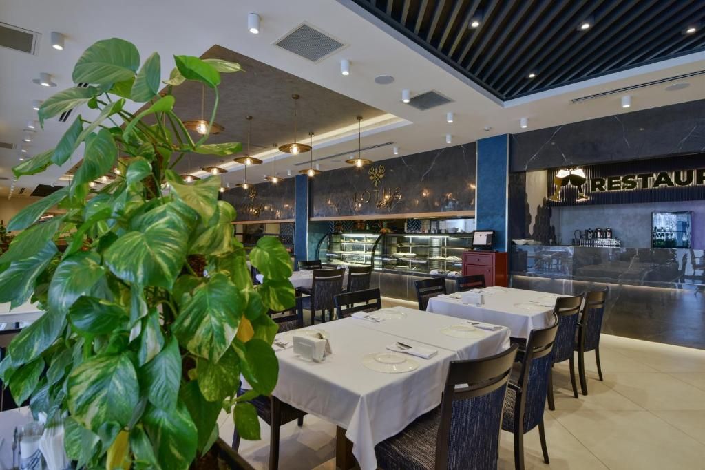 Interior de restaurante com mesas preparadas para refeições, uma grande planta verde em primeiro plano e uma área de balcão com uma placa indicando 'RESTAURANT'.