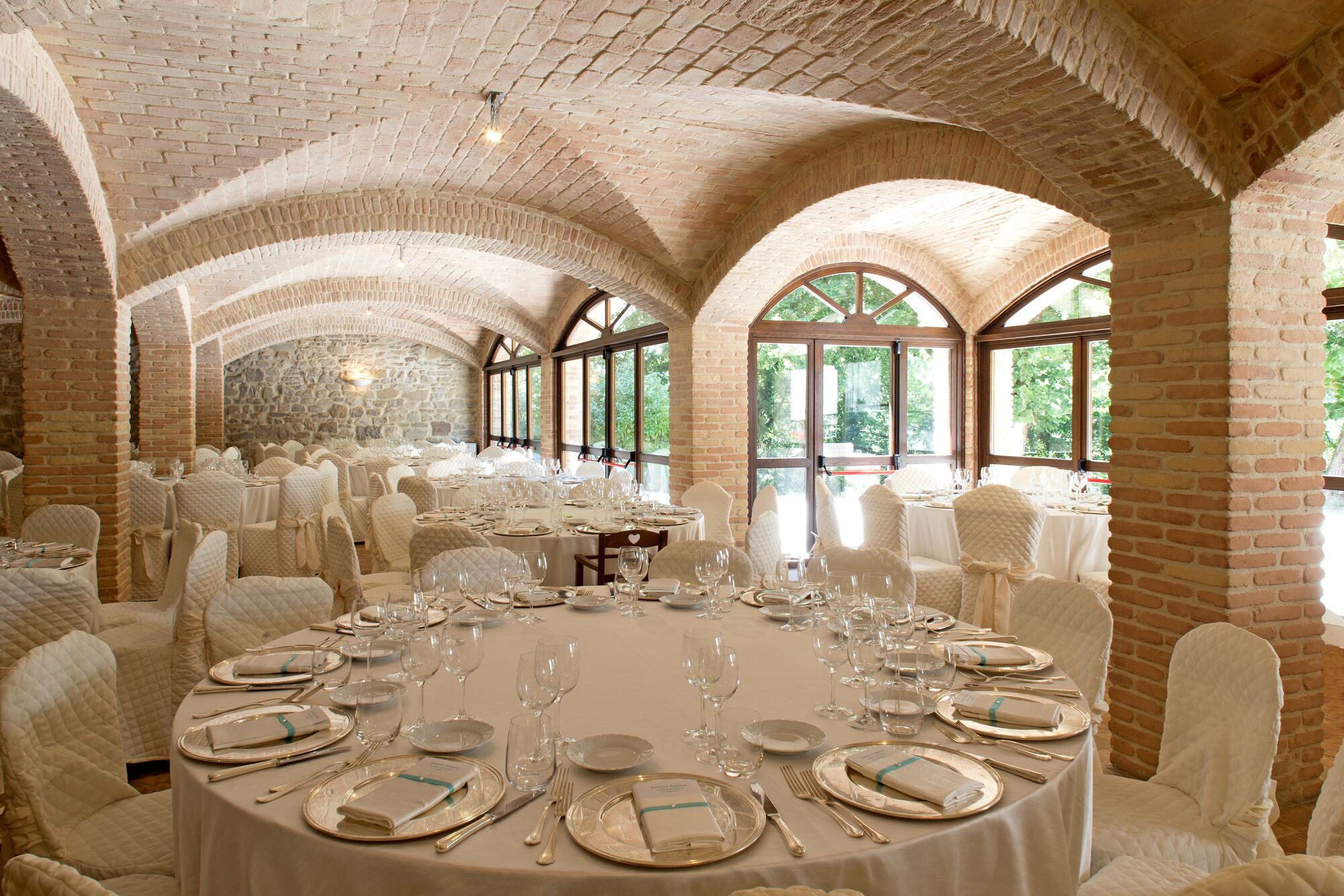 Uma sala de banquetes com mesas redondas preparadas para um evento formal, apresentando arcos de tijolo e grandes janelas.