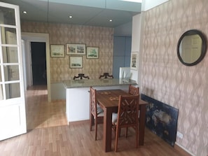 Uma sala de jantar com mesa e cadeiras de madeira, cozinha com bancada e parede decorada com quadros.