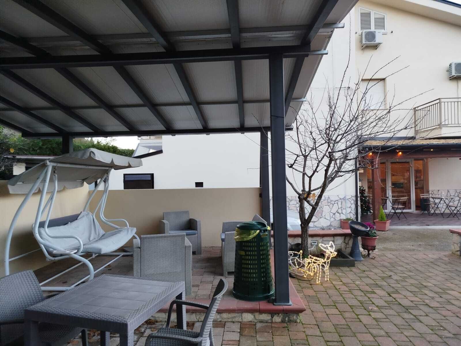 Um pátio externo com área de estar coberta, incluindo cadeiras, mesas, uma lixeira e uma árvore decorativa.