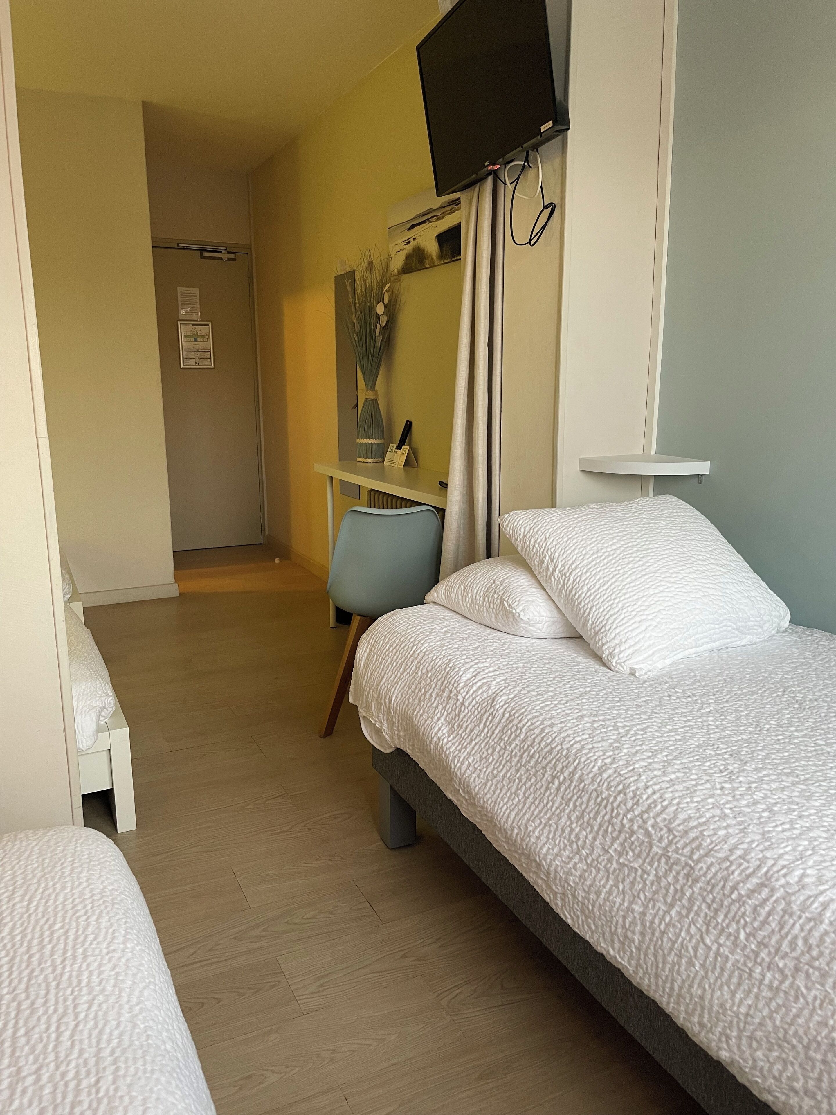 Quarto de hotel com uma cama, uma escrivaninha com cadeira, uma televisão na parede e uma porta ao fundo.