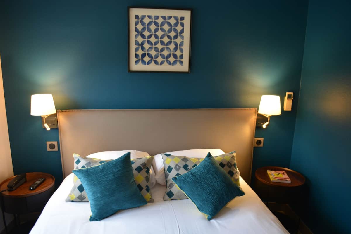 Quarto de hotel com uma cama, duas mesas de cabeceira, uma luminária na parede e uma obra de arte emoldurada.