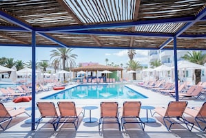 Área da piscina com espreguiçadeiras e guarda-sóis, uma piscina e uma área de estar coberta com mesas.