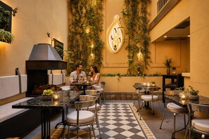Uma área de refeições ao ar livre com piso xadrez, cadeiras e mesas de metal, cercada por vegetação e luminárias decorativas.