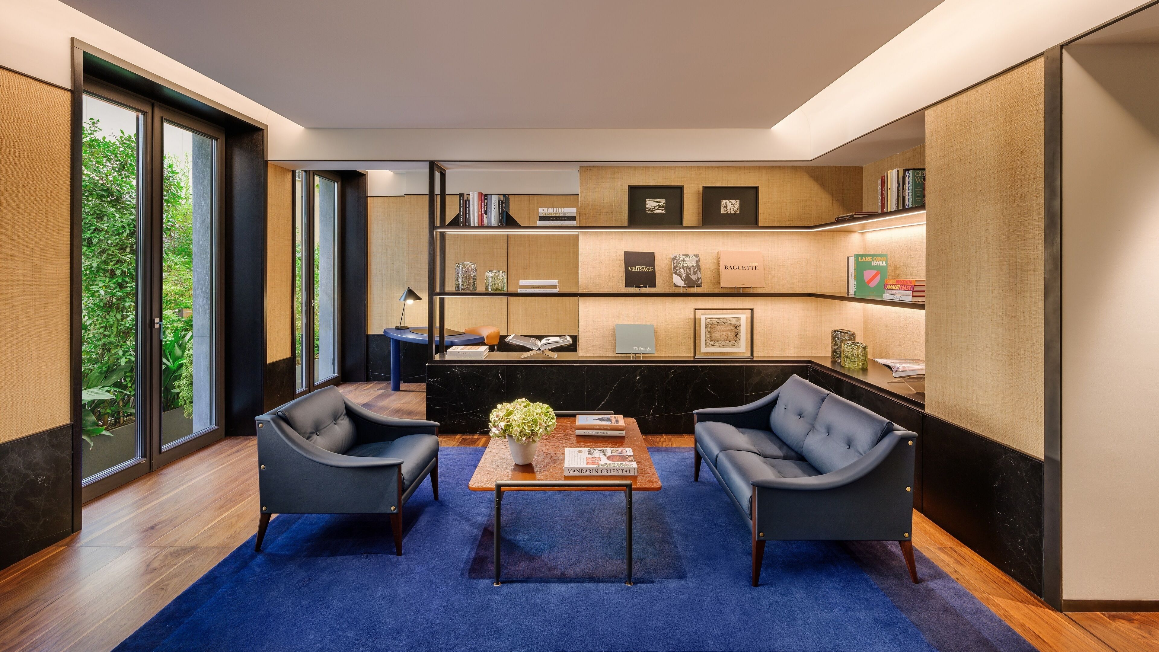 Uma área de recepção moderna com carpete azul, duas poltronas de couro preto, uma mesa de centro de madeira com uma planta e uma estante com vários livros e quadros.