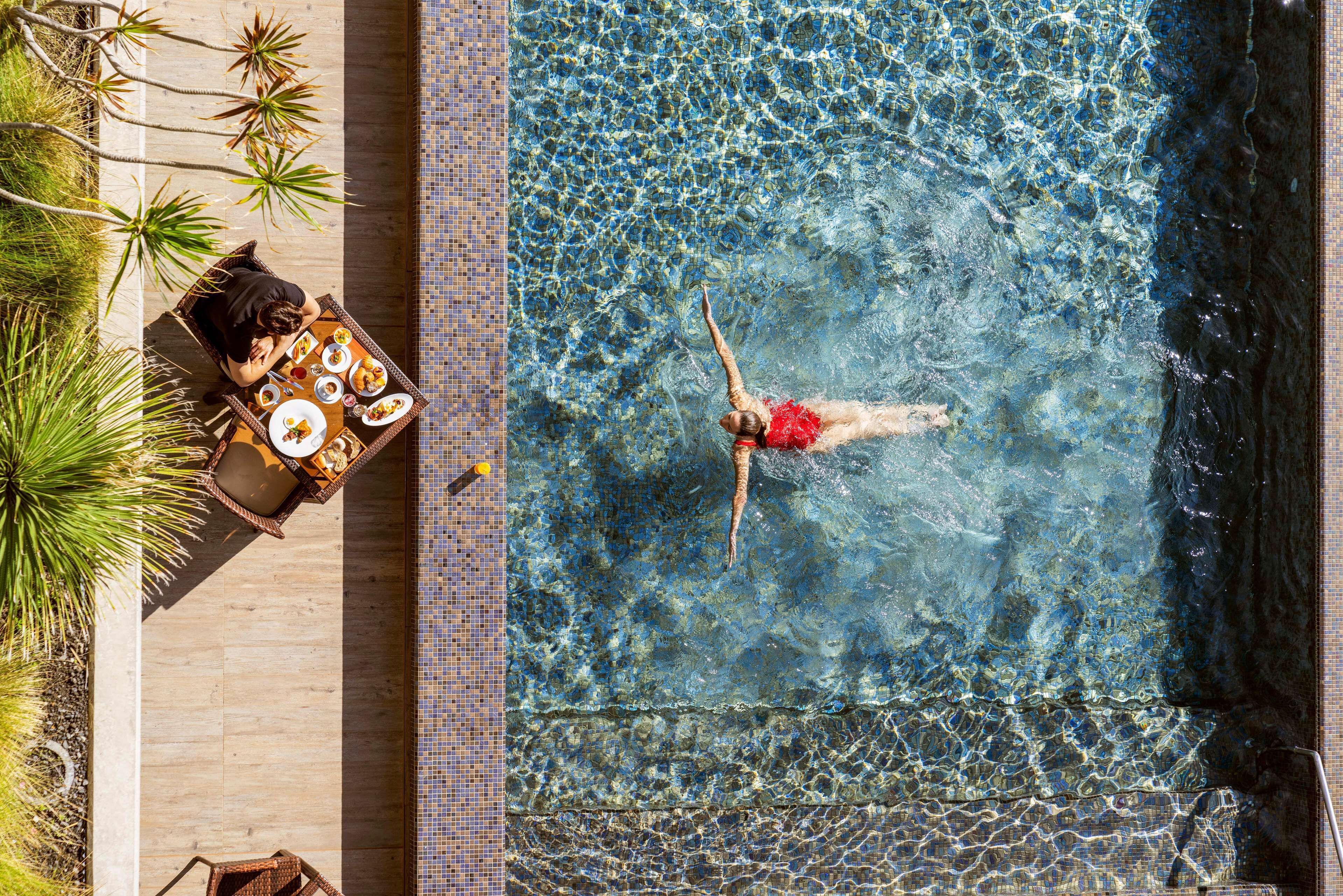 Vista aérea de uma piscina com uma pessoa nadando, área de refeições com mesa e cadeiras, e vegetação ao redor.