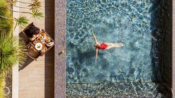 Vista aérea de uma piscina com uma pessoa nadando, área de refeições com mesa e cadeiras, e vegetação ao redor.