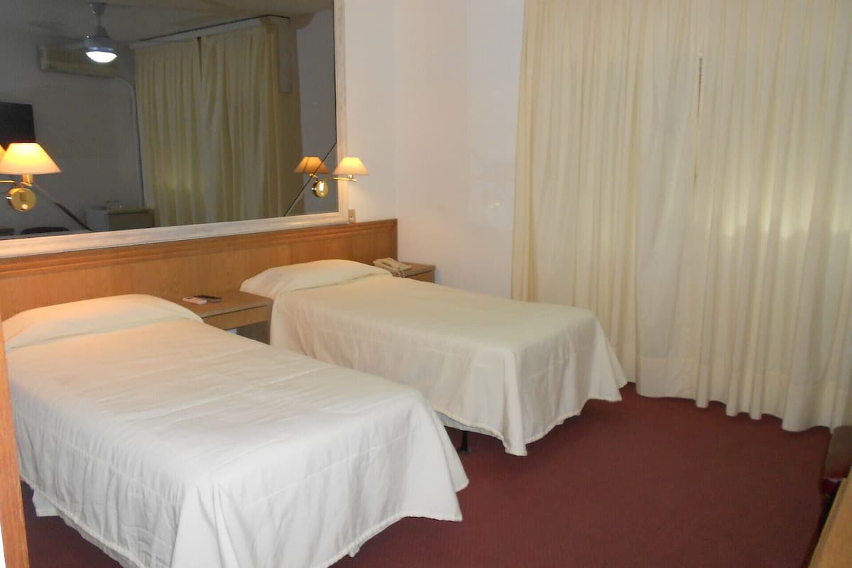 Quarto de hotel com duas camas de solteiro, cabeceira de madeira, mesa de cabeceira com abajur, telefone e um espelho grande.