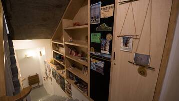 Uma escadaria leva a um quarto com estantes de madeira cheias de livros e revistas, um mural com diversos avisos e uma porta com uma placa pendurada.