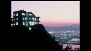 Um edifício alto com janelas iluminadas, com vista para uma paisagem urbana e um corpo de água ao entardecer.