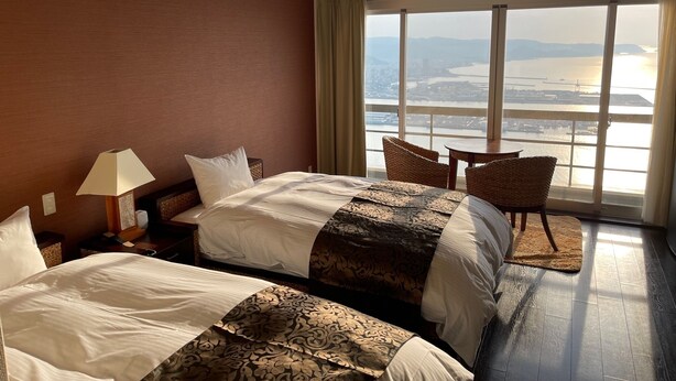 Quarto de hotel com duas camas, uma mesa de cabeceira, uma luminária e vista para um corpo de água e montanhas.