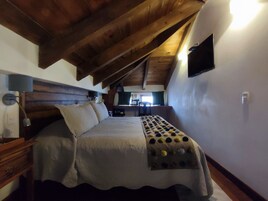 Quarto com teto de madeira, cama com colcha branca, mesa de cabeceira com abajur e uma pequena televisão na parede.