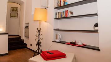 Quarto de hotel com uma cama, uma mesa de cabeceira com toalha vermelha e controle remoto, uma luminária, uma estante com livros, uma pequena prateleira com bule e xícaras, e uma escada que leva a um andar superior.