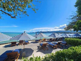 Um resort à beira-mar com guarda-sóis listrados, cadeiras de praia e vista para o oceano e montanhas.