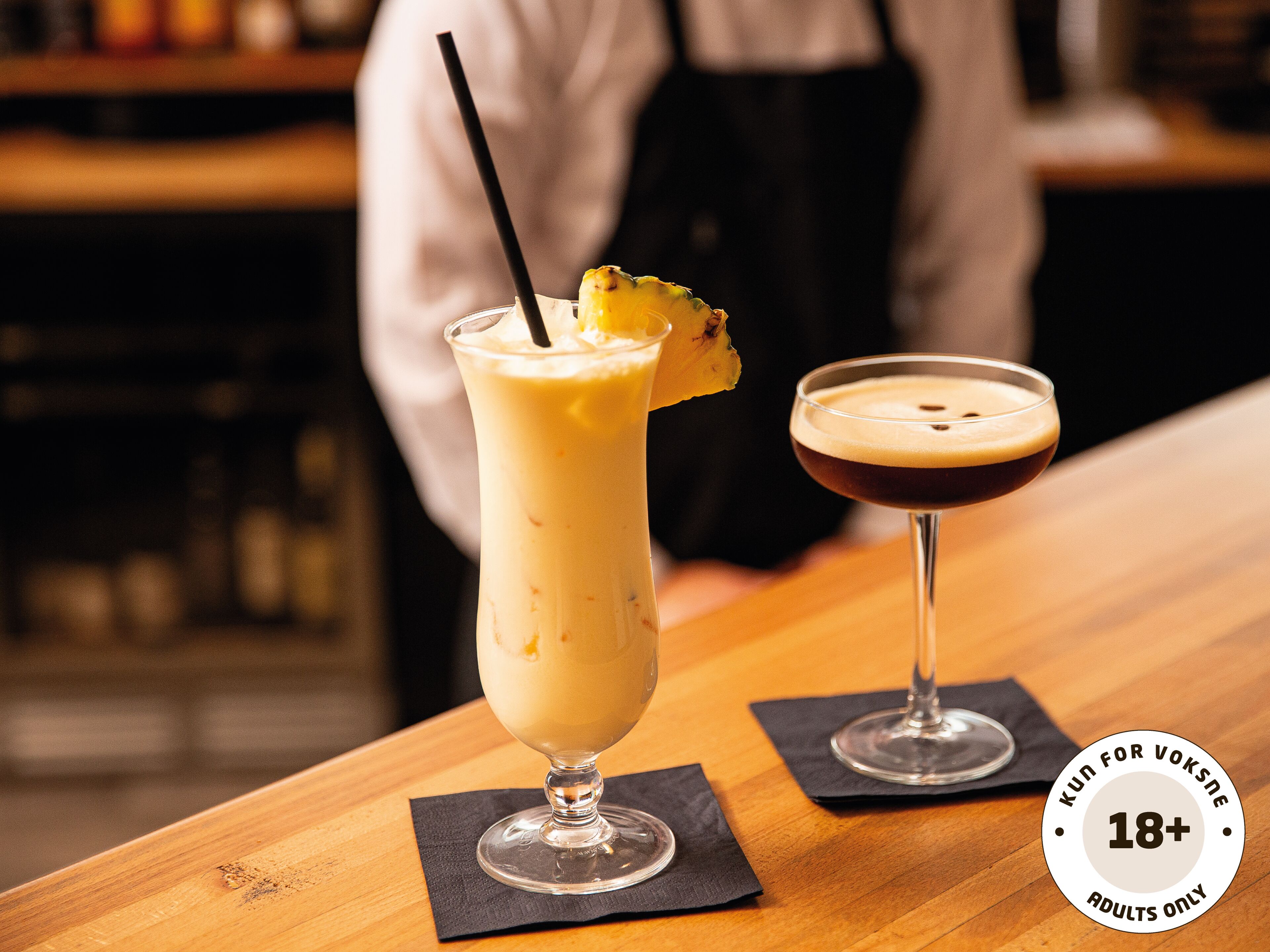 Duas bebidas em um balcão de bar com um aviso: 'KUN FOR VOKSNE 18+'.