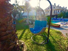 Uma cadeira suspensa com almofada azul em área gramada, com piscina e cadeiras de sol ao fundo.