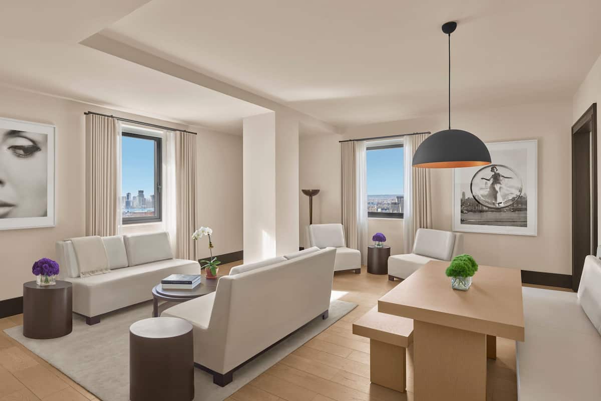 Uma sala de estar moderna com um sofá, poltronas, uma mesa de centro e uma mesa de jantar. Há grandes janelas com cortinas, vista da cidade e obras de arte nas paredes.