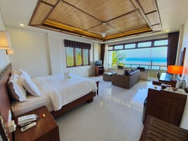 Quarto de hotel com cama grande, sofá, televisão, varanda com vista para o mar e ventoinha no teto.