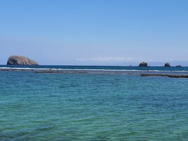 Vista costeira com águas azul-claras, afloramentos rochosos e uma ilha ao longe.
