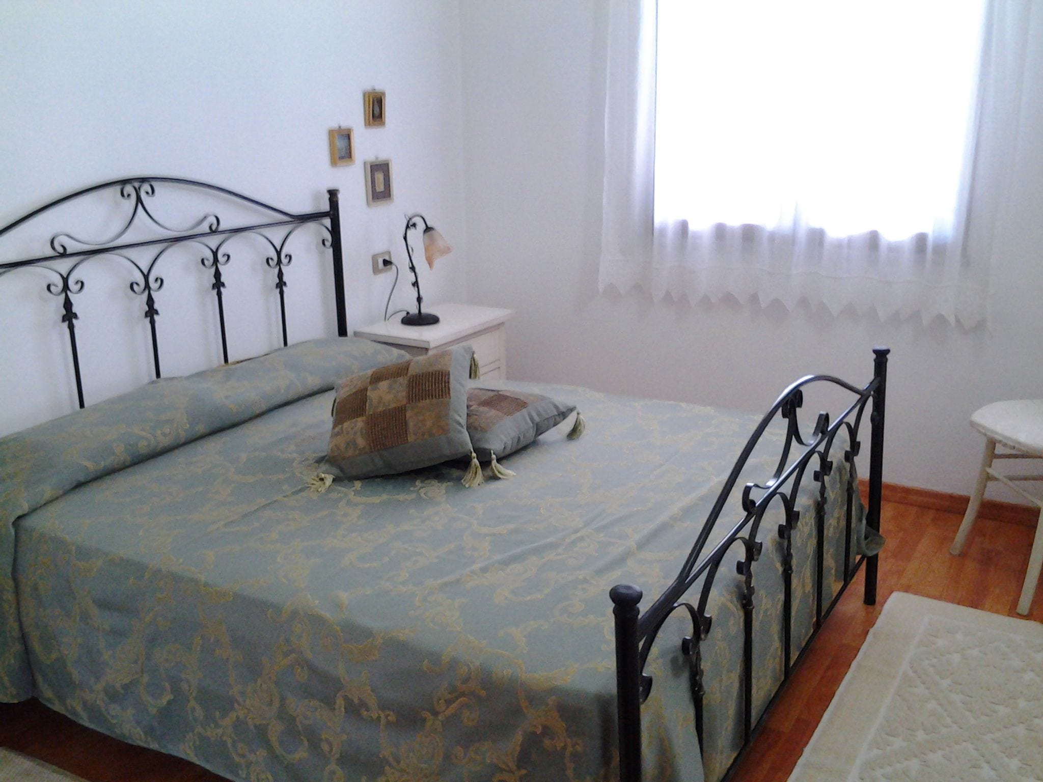 Cama bem arrumada com colcha estampada e almofadas, mesa de cabeceira com abajur e janela com cortina translúcida.