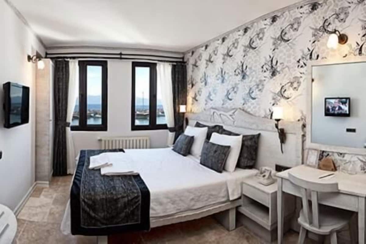Quarto de hotel com cama, escrivaninha, cadeira, televisão e vista para o mar pelas janelas.