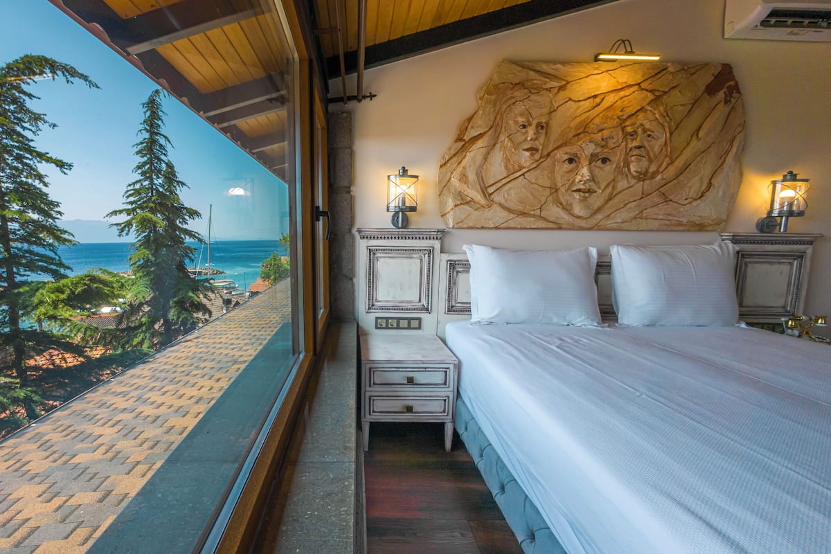 Quarto com uma cama grande, duas mesas de cabeceira, uma obra de arte na parede e vista para o oceano e árvores.