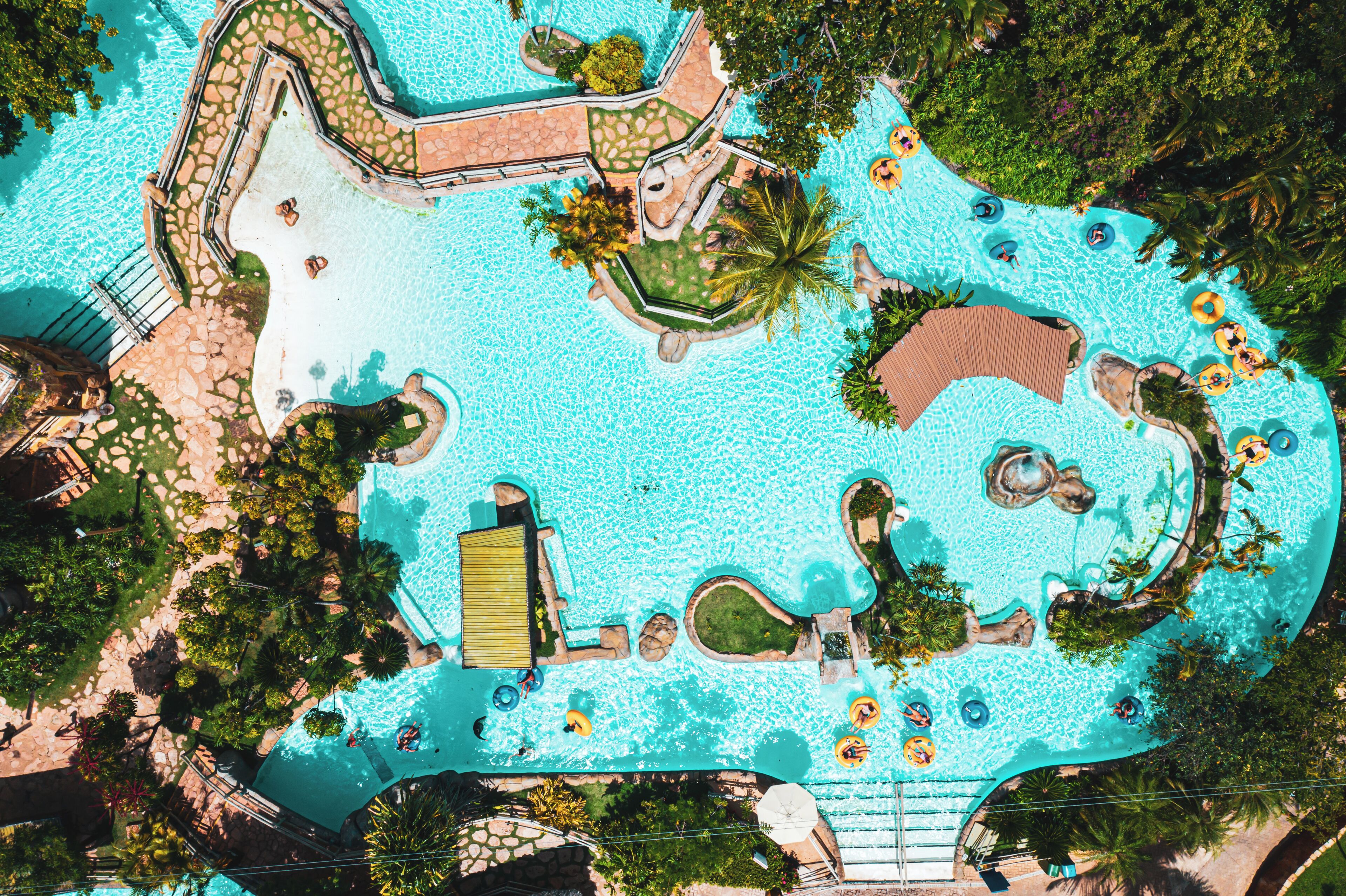 Vista aérea de uma grande piscina circular com várias pistas de natação, rodeada por vegetação exuberante e palmeiras.