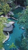 Uma piscina azul límpida rodeada por vegetação exuberante, uma estrutura com telhado de palha e uma pequena ponte.