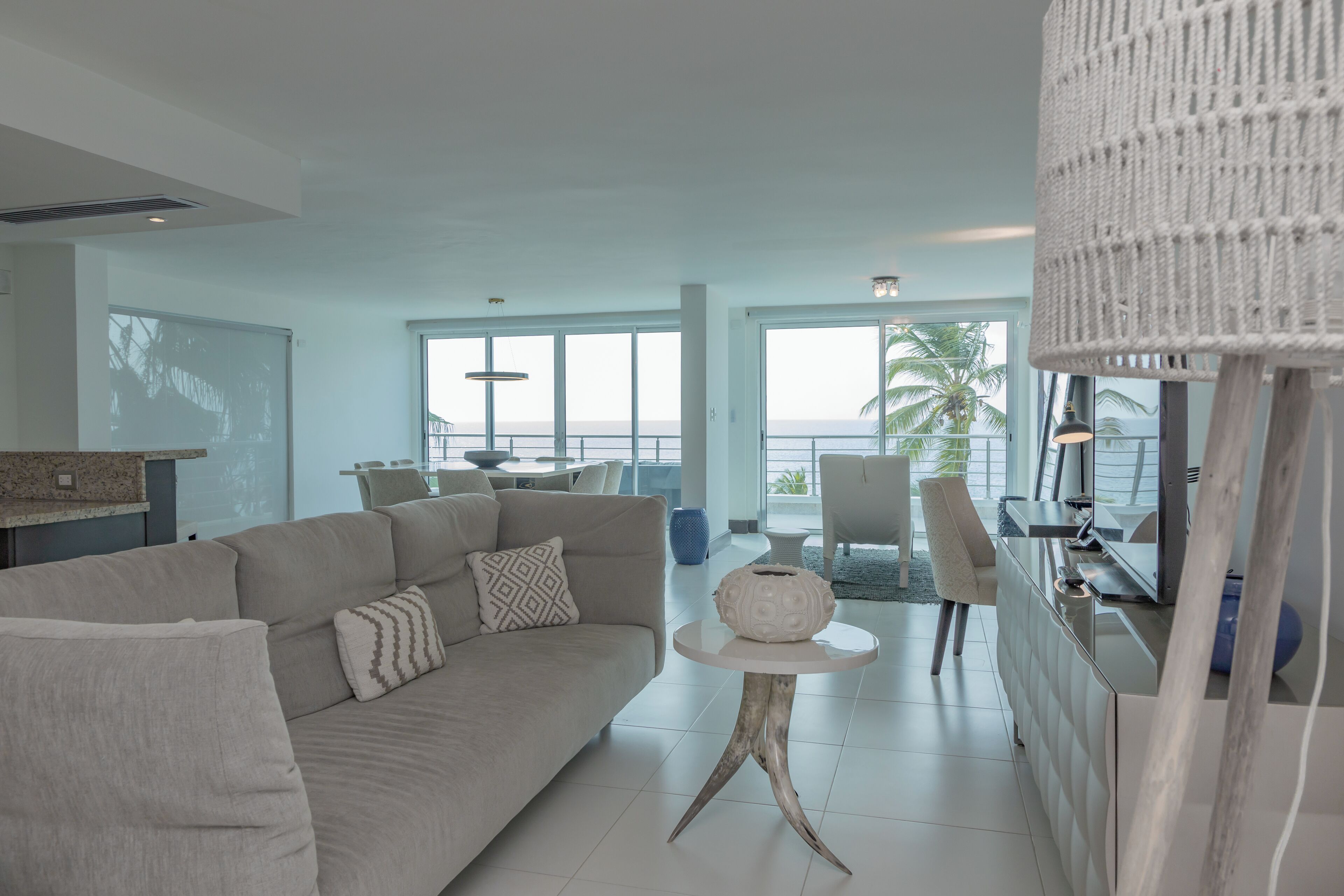 Uma sala de estar moderna com um sofá grande, uma mesa de centro redonda e vista para o oceano através de amplas janelas.