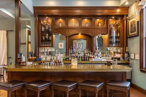 Uma área de bar com bancada de granito polido, diversas garrafas de bebidas e vários tipos de copos.
