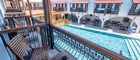 Uma área à beira da piscina com cadeiras e mesas de madeira, piscina e um edifício com varandas e assentos externos.