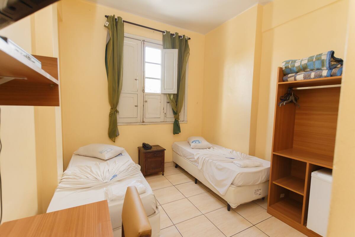 Quarto pequeno e limpo com duas camas de solteiro, uma escrivaninha de madeira, uma cadeira, um criado-mudo e um guarda-roupa.