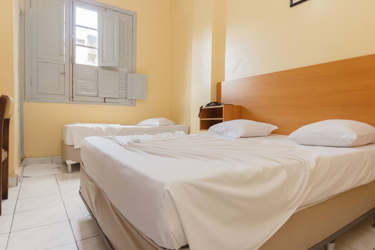 Quarto de hotel com uma cama grande, uma mesa de cabeceira, um guarda-roupa e uma janela com persianas.
