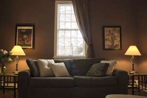 Uma sala de estar com sofá, duas luminárias, janela com cortinas e duas imagens emolduradas na parede.