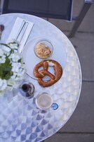 Mesa redonda com um prato branco contendo um pretzel e uma pequena tigela de molho, uma garrafa de molho e um copo de cerveja.