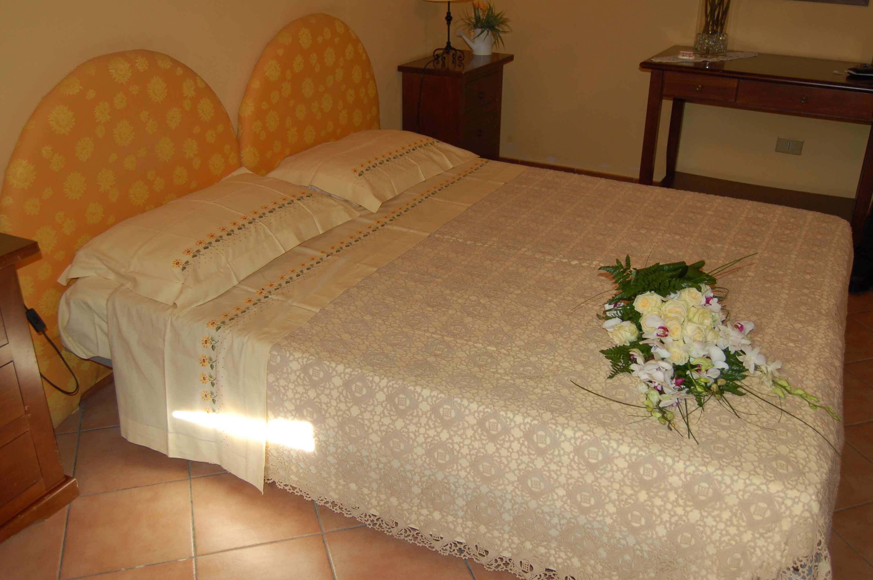 Cama bem arrumada com cabeceira estampada em floral, colcha com detalhes em renda e um buquê de flores no lado direito.
