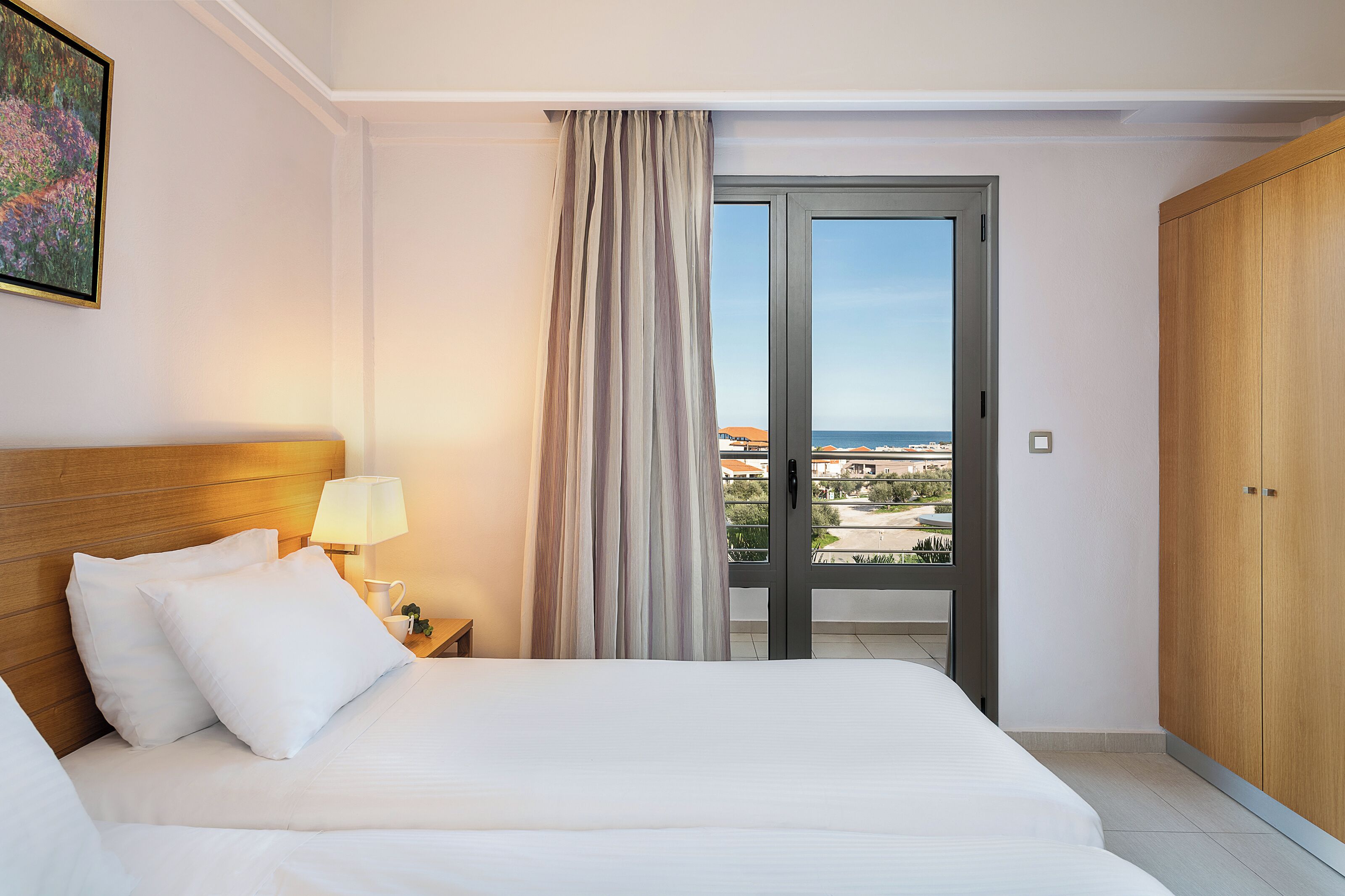 Quarto de hotel com cama, mesa de cabeceira, luminária, guarda-roupa e vista da cidade através de uma grande janela.