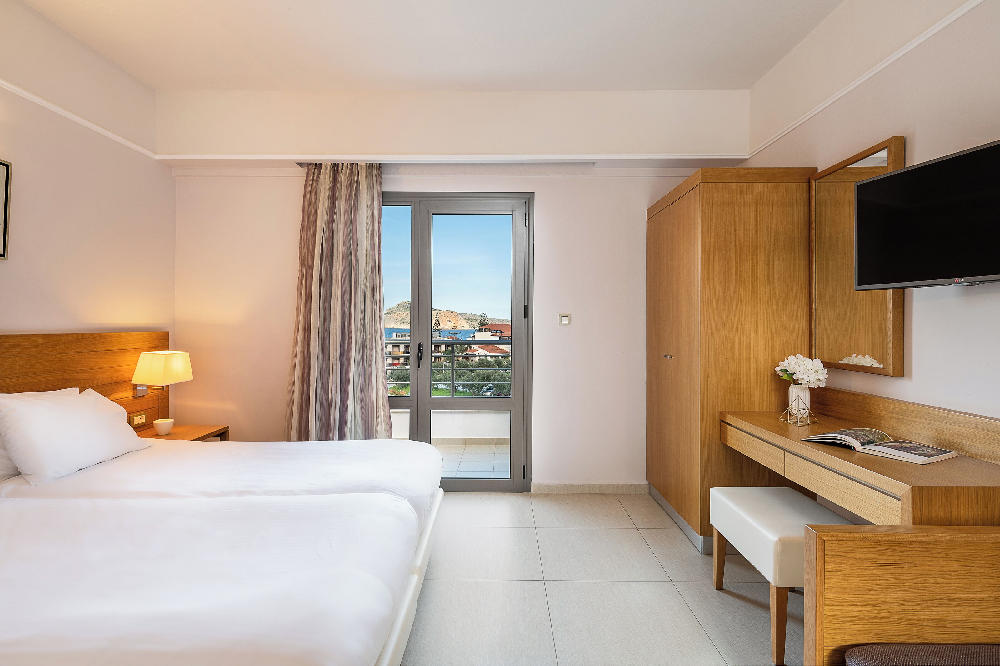 Quarto de hotel com uma cama, uma escrivaninha, uma televisão e vista para um edifício através de uma porta de vidro.