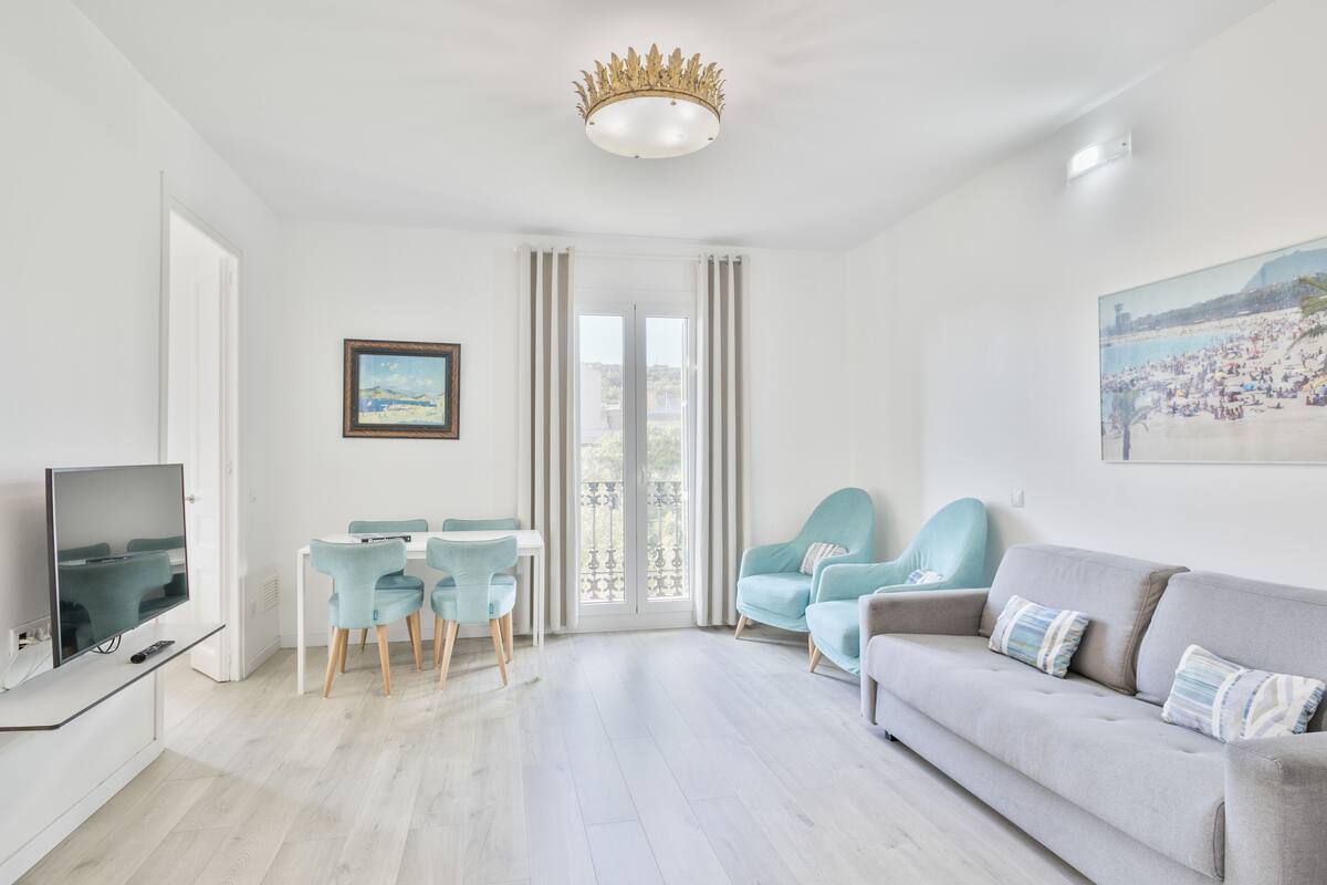 Uma sala de estar moderna com televisão de tela plana, uma área de jantar com cadeiras, um sofá e uma janela grande com cortinas.